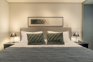 Cama bem arrumada com cobertura acolchoada, duas luminárias de cabeceira e uma obra de arte emoldurada acima da cabeceira.