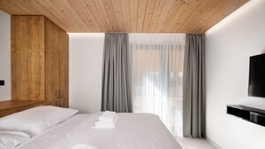 Quarto moderno com teto de madeira, uma cama grande com roupa de cama branca, cortinas cinzas e uma TV na parede.