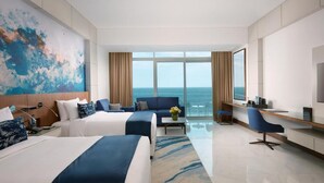 Quarto de hotel com duas camas, uma escrivaninha, uma cadeira, televisão e uma janela grande com vista para o mar.