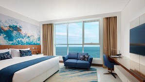 Quarto de hotel com cama, sofá, escrivaninha, televisão e uma grande janela vista para o mar.