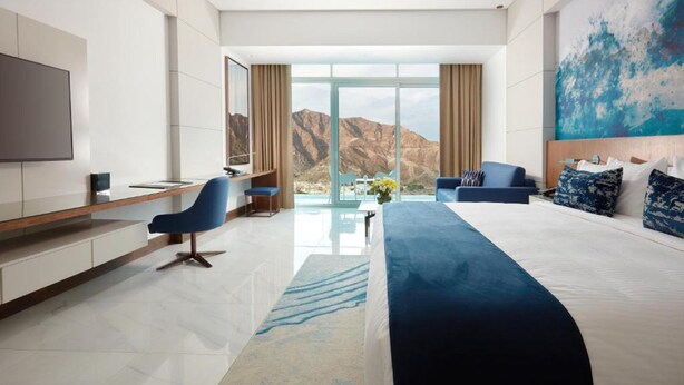 Quarto de hotel moderno com uma cama grande, uma escrivaninha, uma cadeira, uma televisão e vista para montanhas.