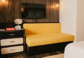 Quarto de hotel moderno com televisão de ecrã plano fixada numa parede de madeira, uma mesa de cabeceira com uma lâmpada e um sofá amarelo.
