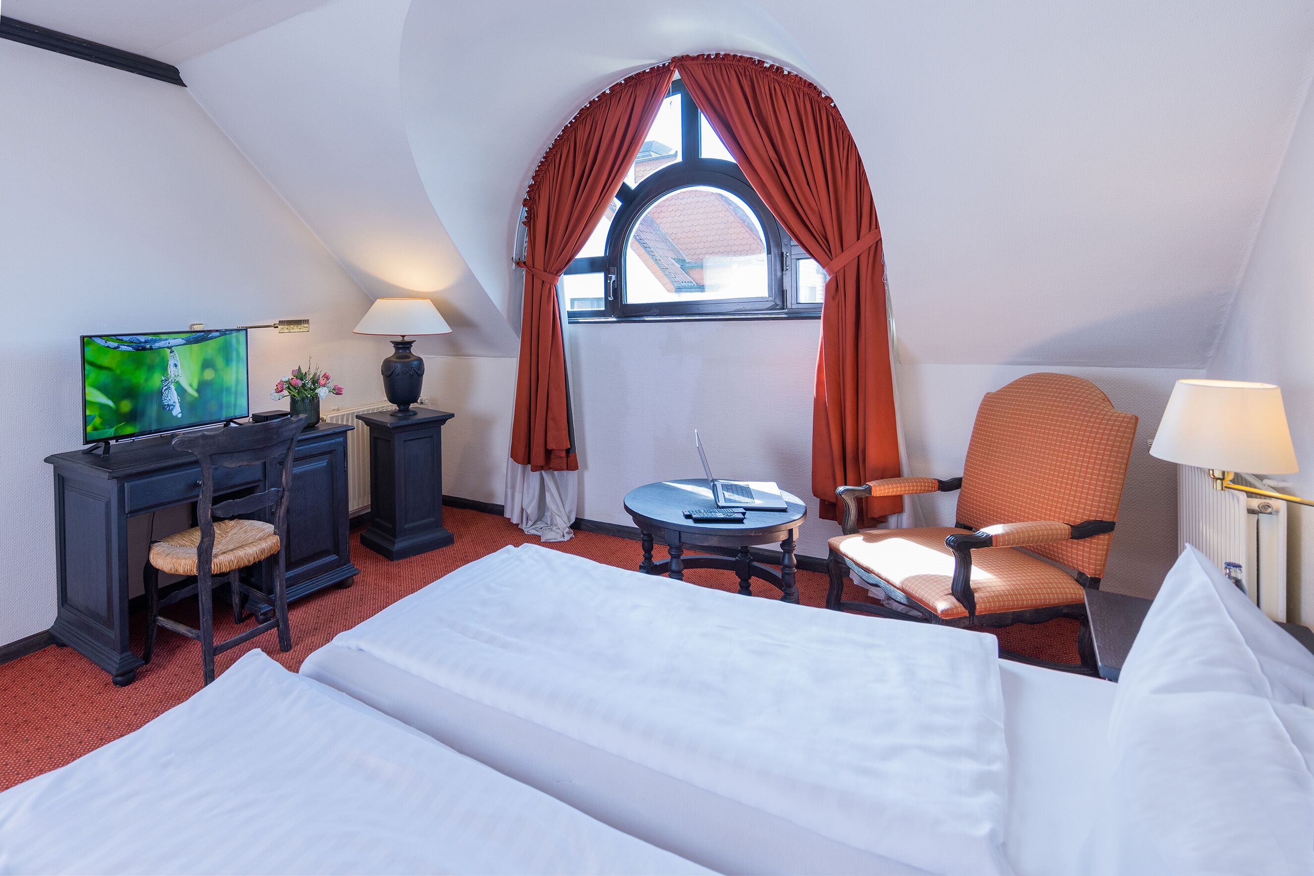 Quarto de hotel com cama, escrivaninha com televisão, cadeira, mesinha e janela com cortinas.