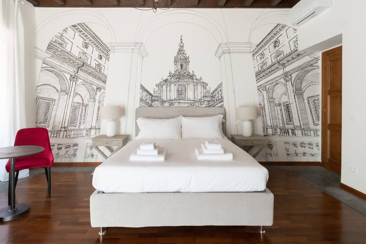 Quarto de hotel com uma cama grande, duas mesas de cabeceira com luminárias, uma cadeira vermelha e um mural na parede com desenhos arquitetônicos.