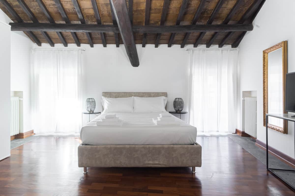 Um quarto espaçoso com uma cama grande, vigas de madeira no teto, cortinas brancas e uma televisão em um móvel.