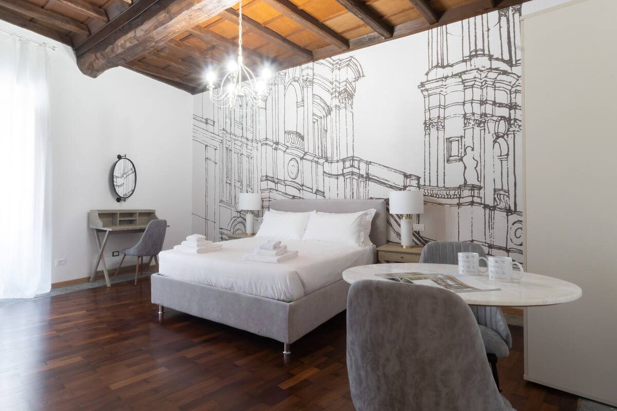 Quarto de hotel moderno com uma cama, escrivaninha, cadeira, mesa redonda e parede com desenhos arquitetônicos.