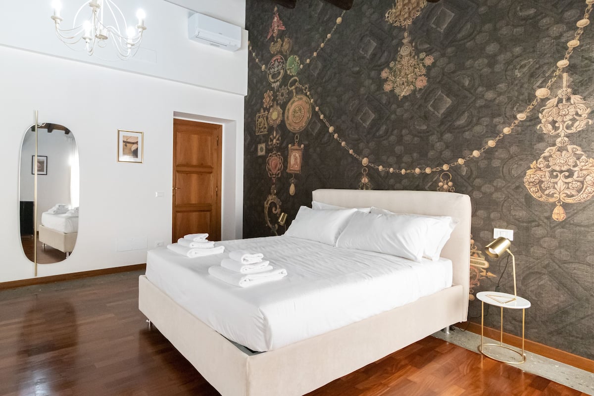 Quarto de hotel com uma cama grande, um espelho, um lustre e um detalhe decorativo na parede.