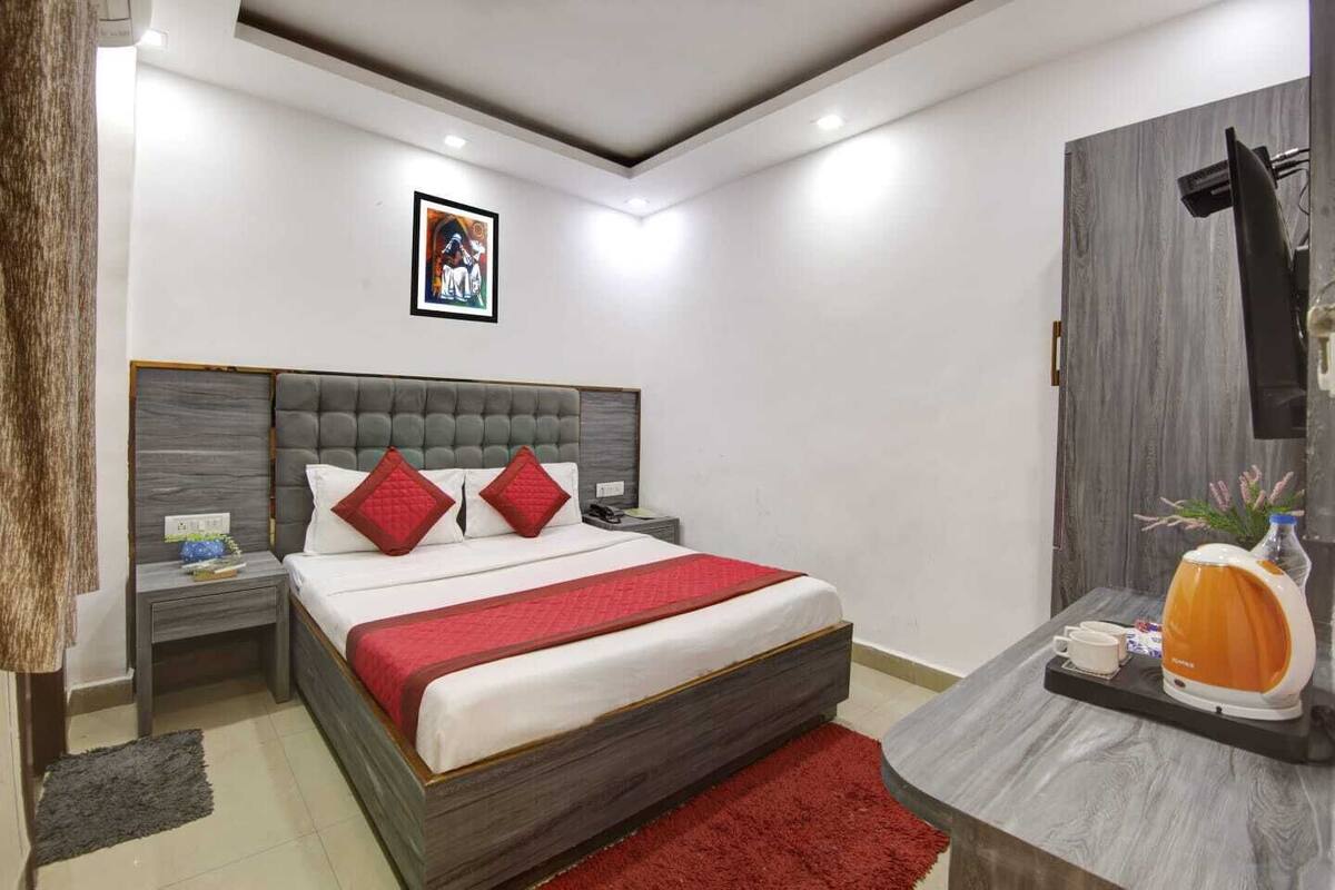 Quarto de hotel moderno com uma cama grande, uma mesa de cabeceira, uma escrivaninha com chaleira e uma TV na parede.