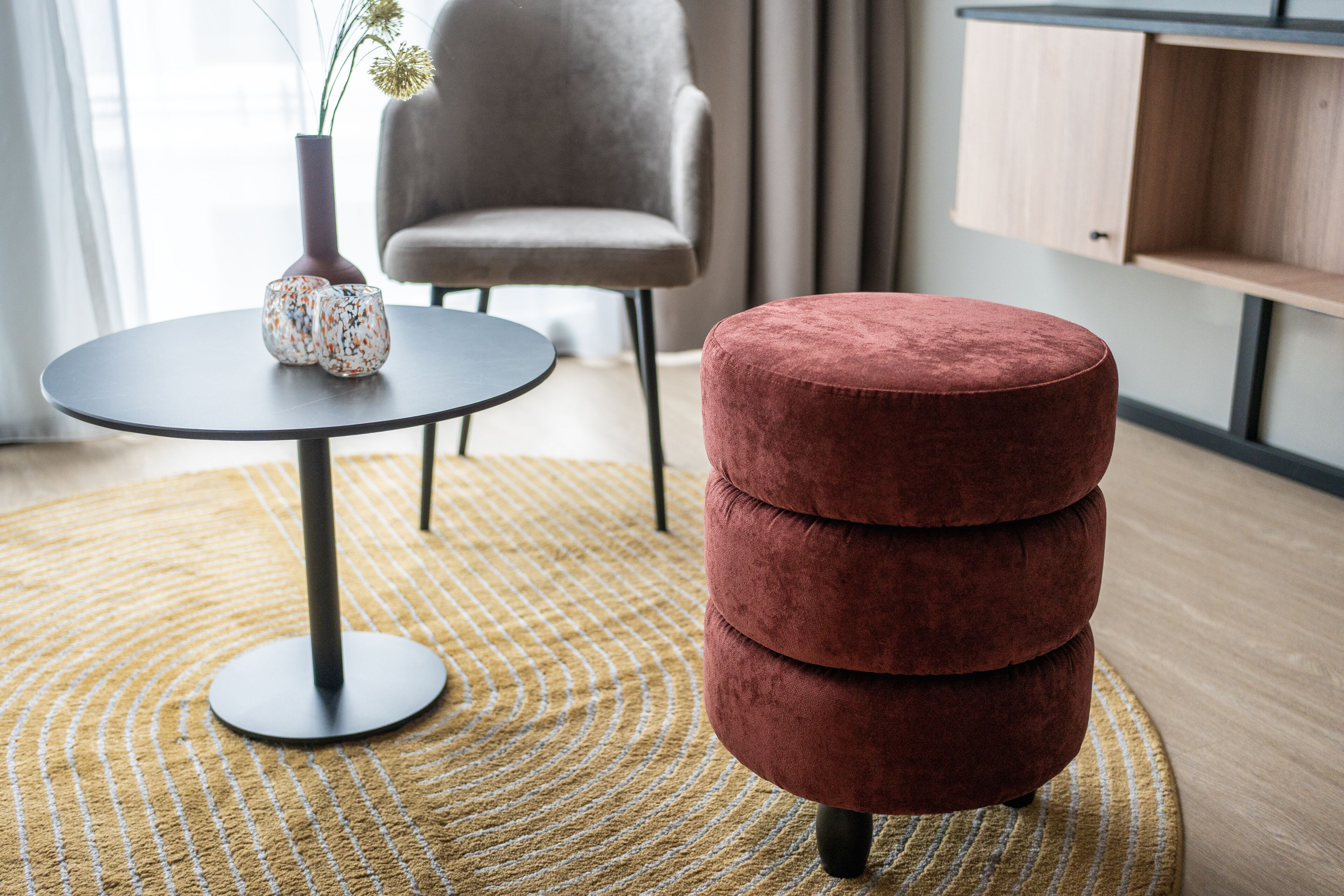 Quarto moderno com uma mesa redonda, um banquinho vermelho, uma cadeira cinza e um vaso com flores.