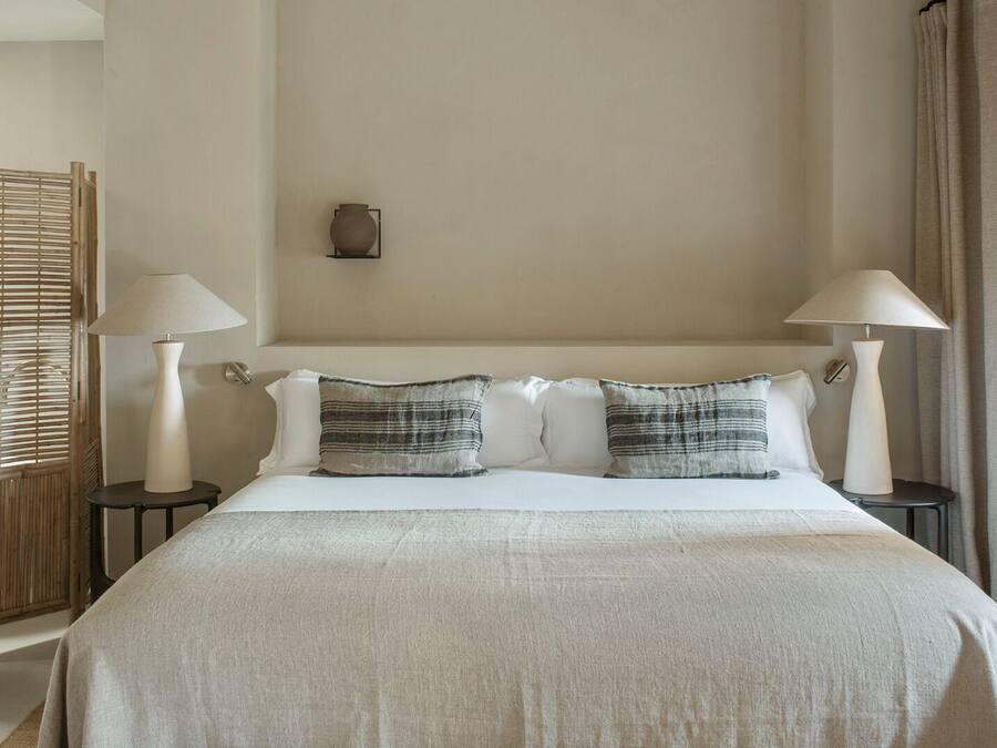 Quarto de hotel com uma cama, duas mesas de cabeceira, duas luminárias, uma luminária na parede e uma janela grande com cortinas.