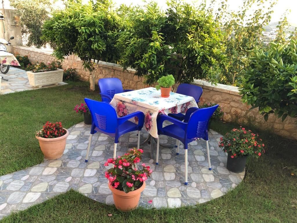 Uma área externa com mesas e cadeiras, um pátio com piso de pedra, uma mesa com toalha de mesa florida e cadeiras azuis.