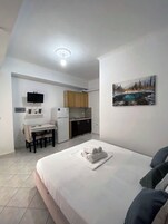 Quarto com cama, pequeno espaço de cozinha, área de refeições e TV na parede.