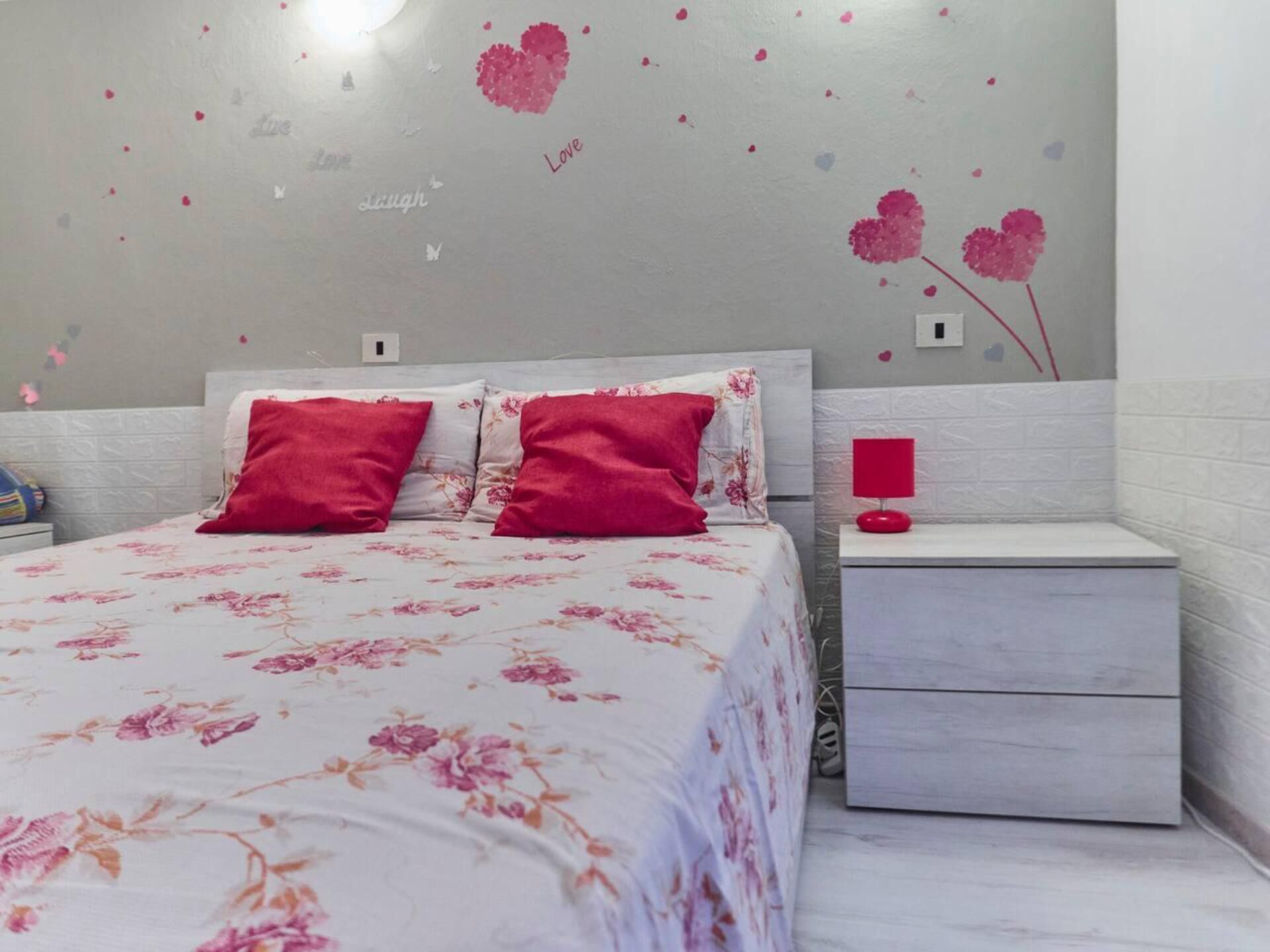 Quarto com cama, duas almofadas, mesa de cabeceira e parede decorada com corações.