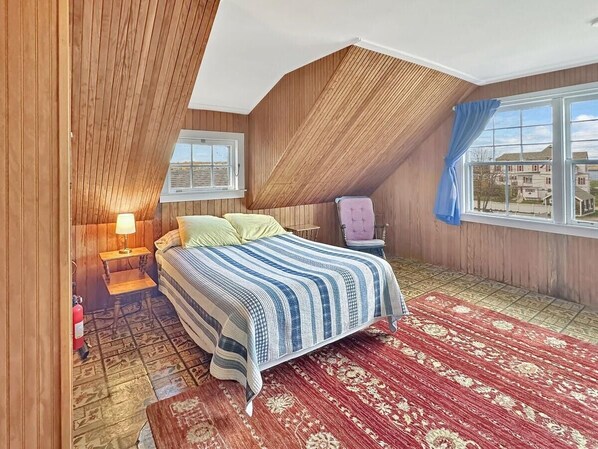 Um quarto com teto inclinado, uma cama com colcha listrada, um tapete vermelho estampado, uma mesa de cabeceira com abajur e uma janela com cortinas.