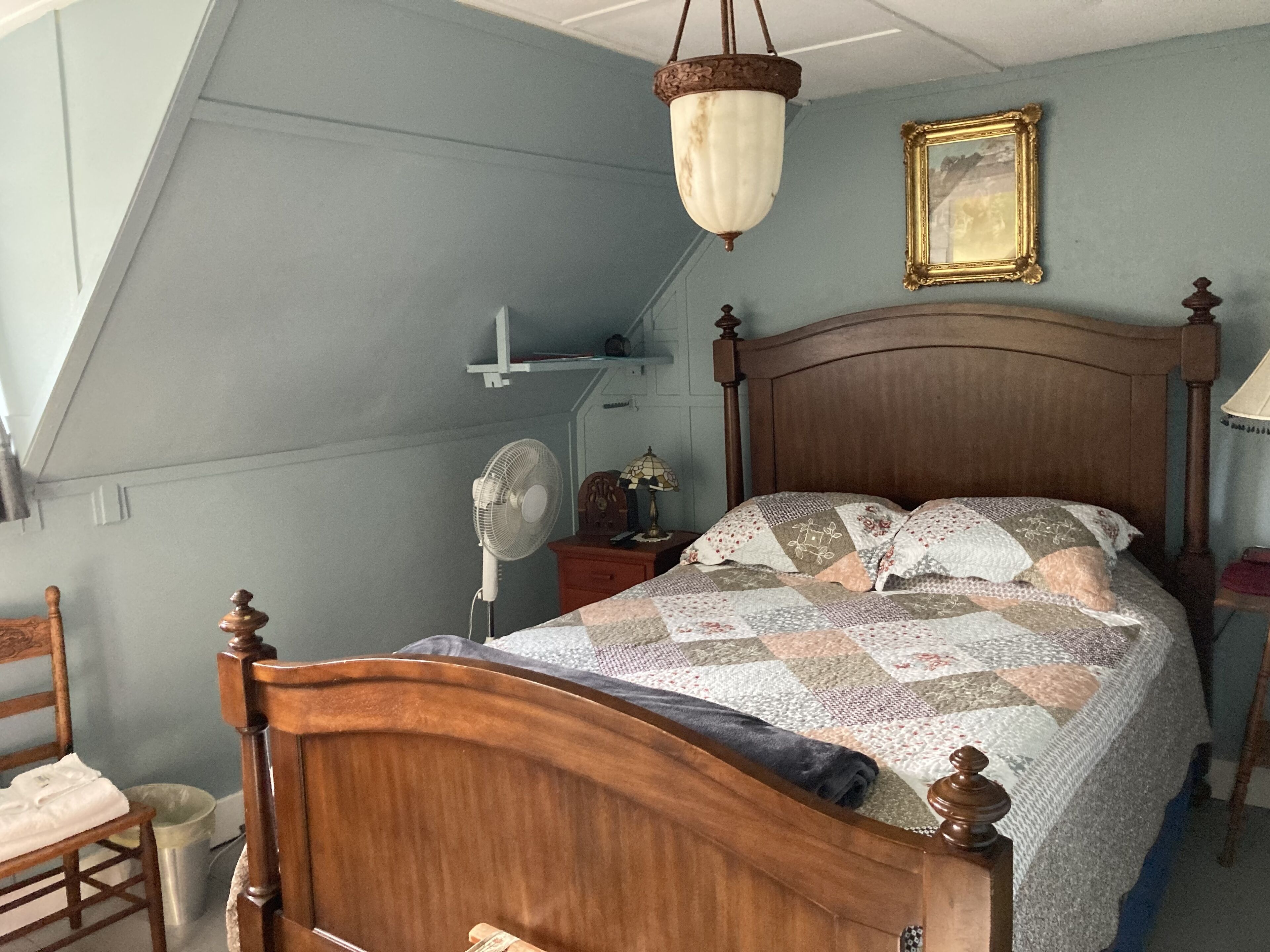 Quarto com cama de madeira, ventilador de teto, abajur e quadro pendurado na parede.