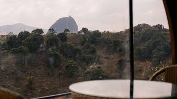 Vista da janela com uma mesa e cadeiras, sobre uma paisagem acidentada com árvores e uma colina imponente ao longe.