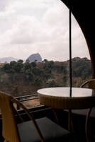 Vista da janela com uma mesa e cadeiras, sobre uma paisagem acidentada com árvores e uma colina imponente ao longe.