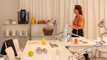 Quarto de hotel moderno com mesa de jantar, cadeiras, máquina de café, cesta de frutas e escrivaninha com abajur.