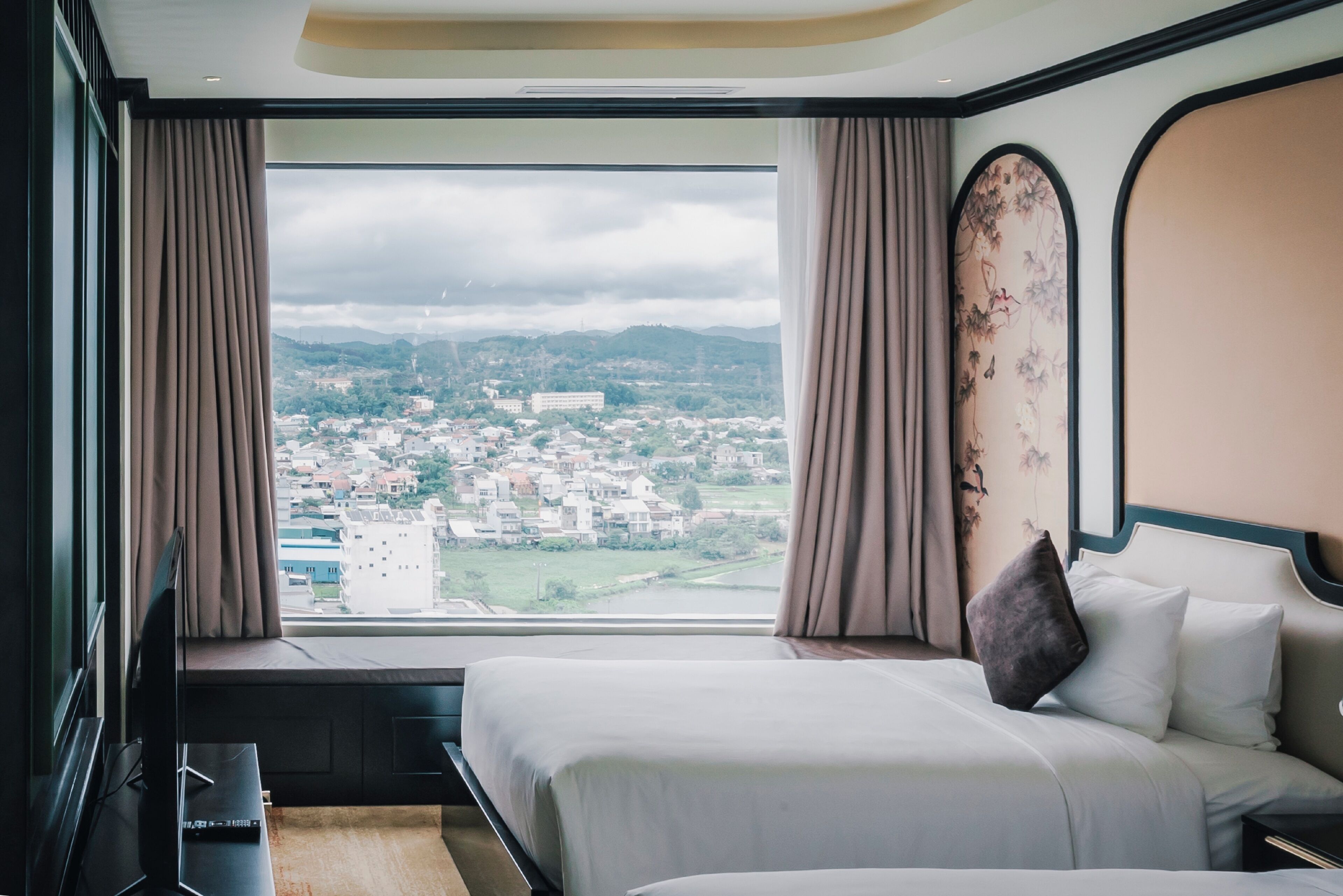 Quarto de hotel com uma janela grande, uma cama, uma mesinha de cabeceira e uma televisão presa à parede.