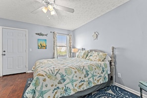 Quarto com cama, ventilador de teto, janela com cortinas e um quadro na parede.