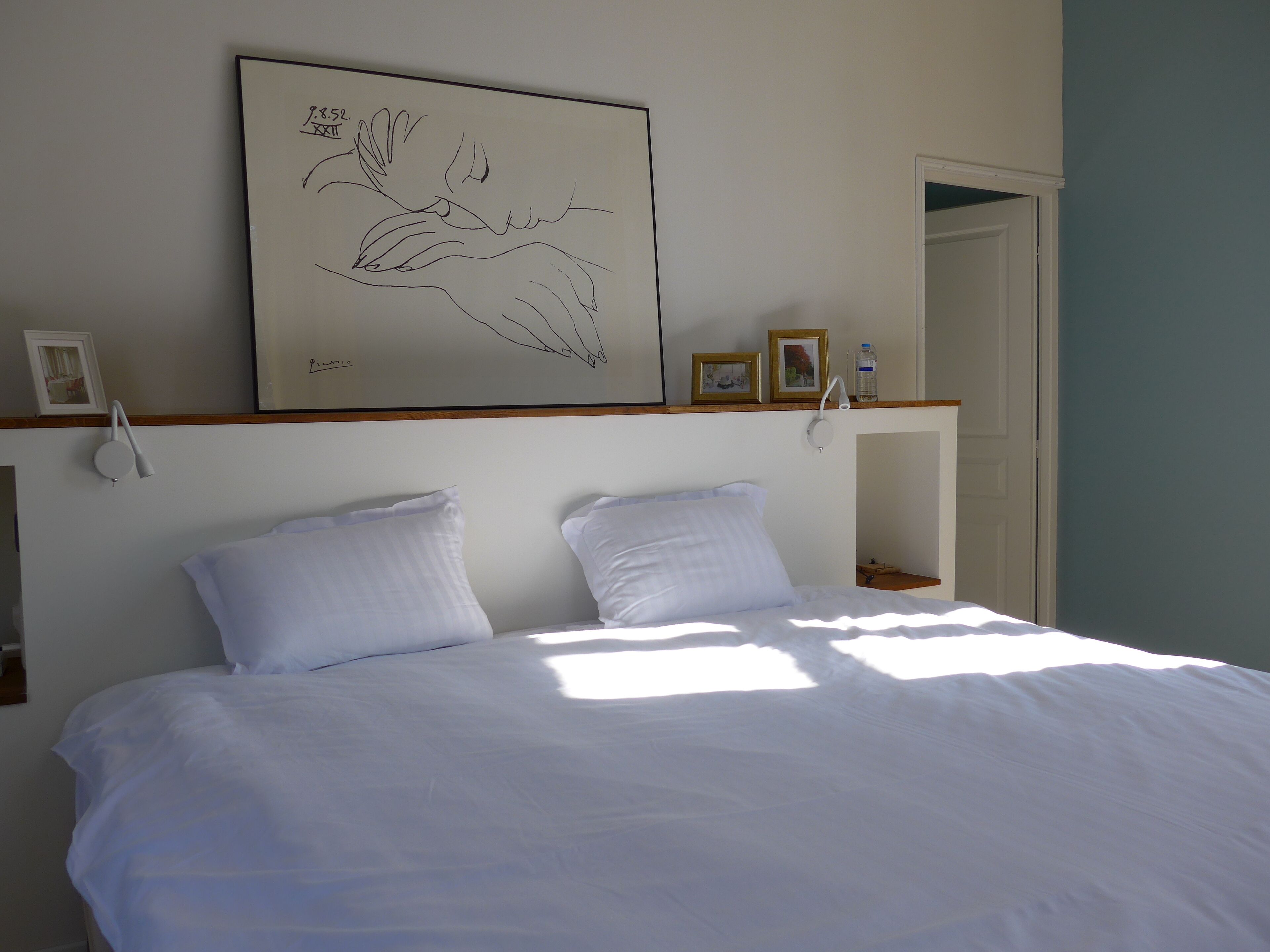 Cama bem arrumada com lençóis brancos, uma obra de arte na parede e uma pequena estante com quadros e uma garrafa.