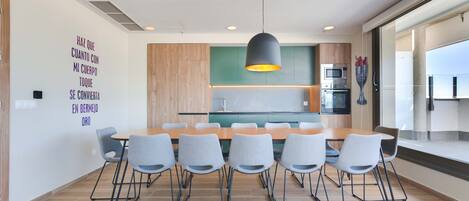 Uma sala de jantar moderna com uma mesa comprida e cadeiras, uma cozinha ao fundo e uma parede decorativa com texto.