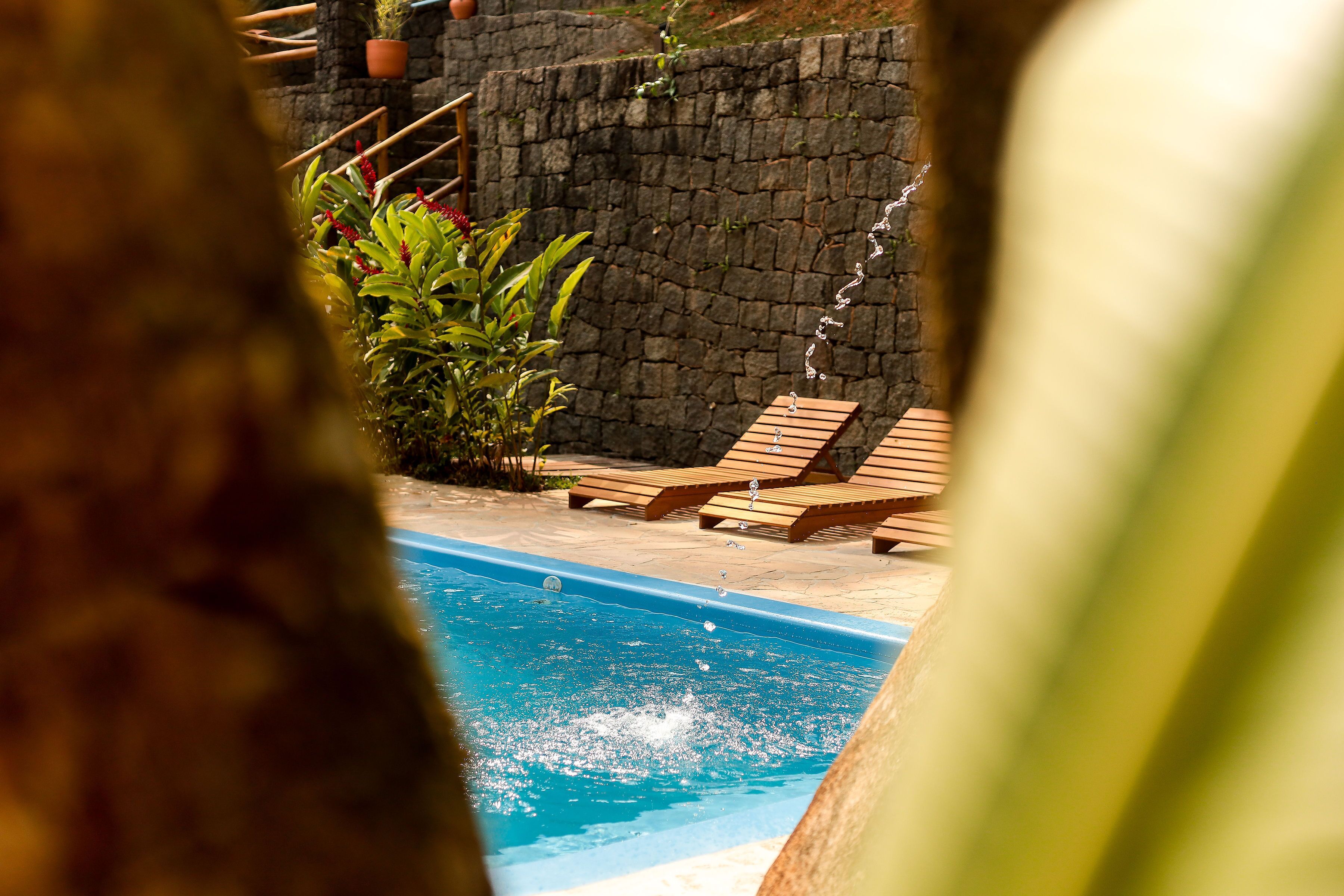 Piscina com espreguiçadeiras de madeira e parede de pedra ao fundo.