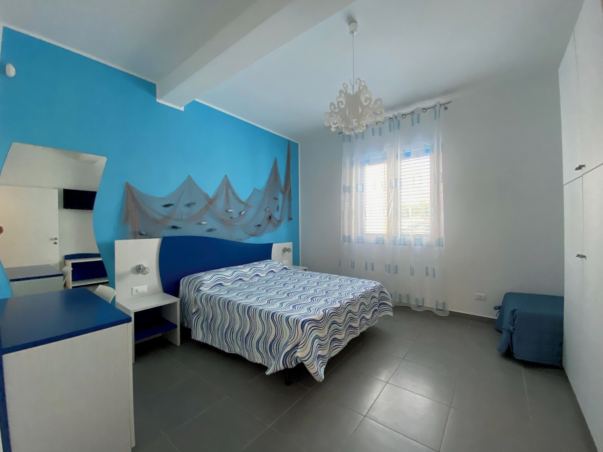 Um quarto com cama, escrivaninha, cadeira, espelho, lustre e janela com cortinas.