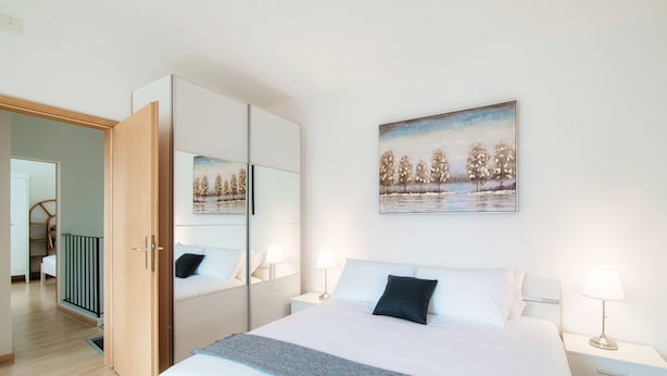 Quarto moderno com uma cama grande, uma mesa de cabeceira com abajur, um criado-mudo e um guarda-roupa, com um quadro na parede.