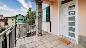 Um edifício residencial com um pilar de pedra, varanda com grade de metal, porta branca com janela e uma pequena área de jardim com um banco.