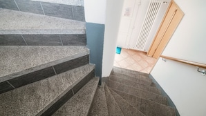 Uma escadaria com degraus alternados em tons escuros e claros, que leva a um quarto com um radiador e uma porta de madeira.