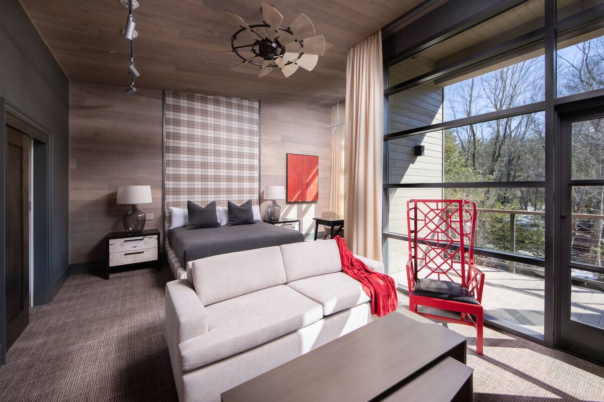Quarto moderno com uma cama grande, um sofá, uma cadeira vermelha e uma varanda com vista para árvores.