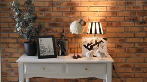 Mesa branca com planta em vaso, quadro emoldurado, luminária e objetos decorativos, tudo em frente a parede de tijolos com uma obra de arte emoldurada acima.