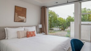 Quarto de hotel com uma cama grande, uma escrivaninha, uma cadeira, uma janela com vista para árvores e um quadro na parede.