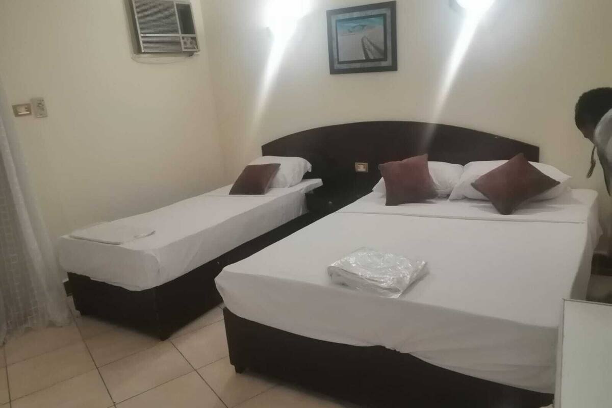 Quarto de hotel com duas camas de solteiro, cabeceira, janela com persianas, um quadro na parede e cortina.