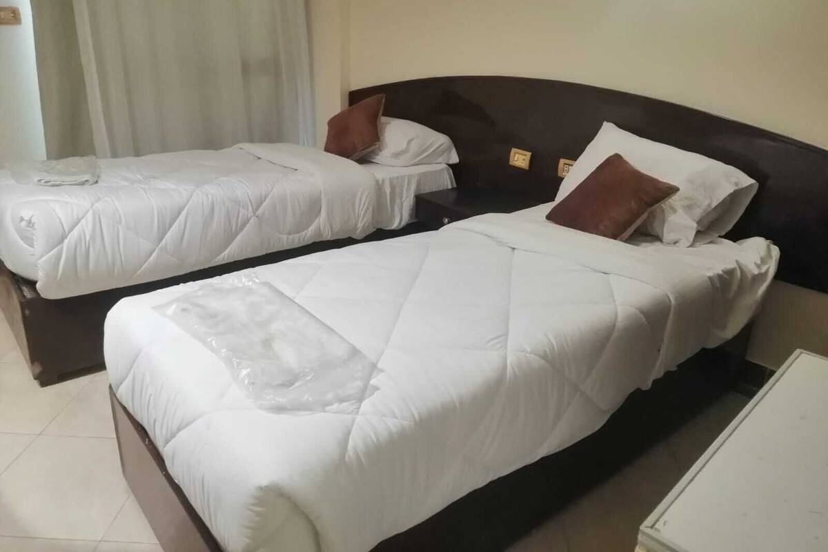 Duas camas de solteiro com roupa de cama branca e almofadas castanhas, um cabeceira escura e uma pequena mesa branca.