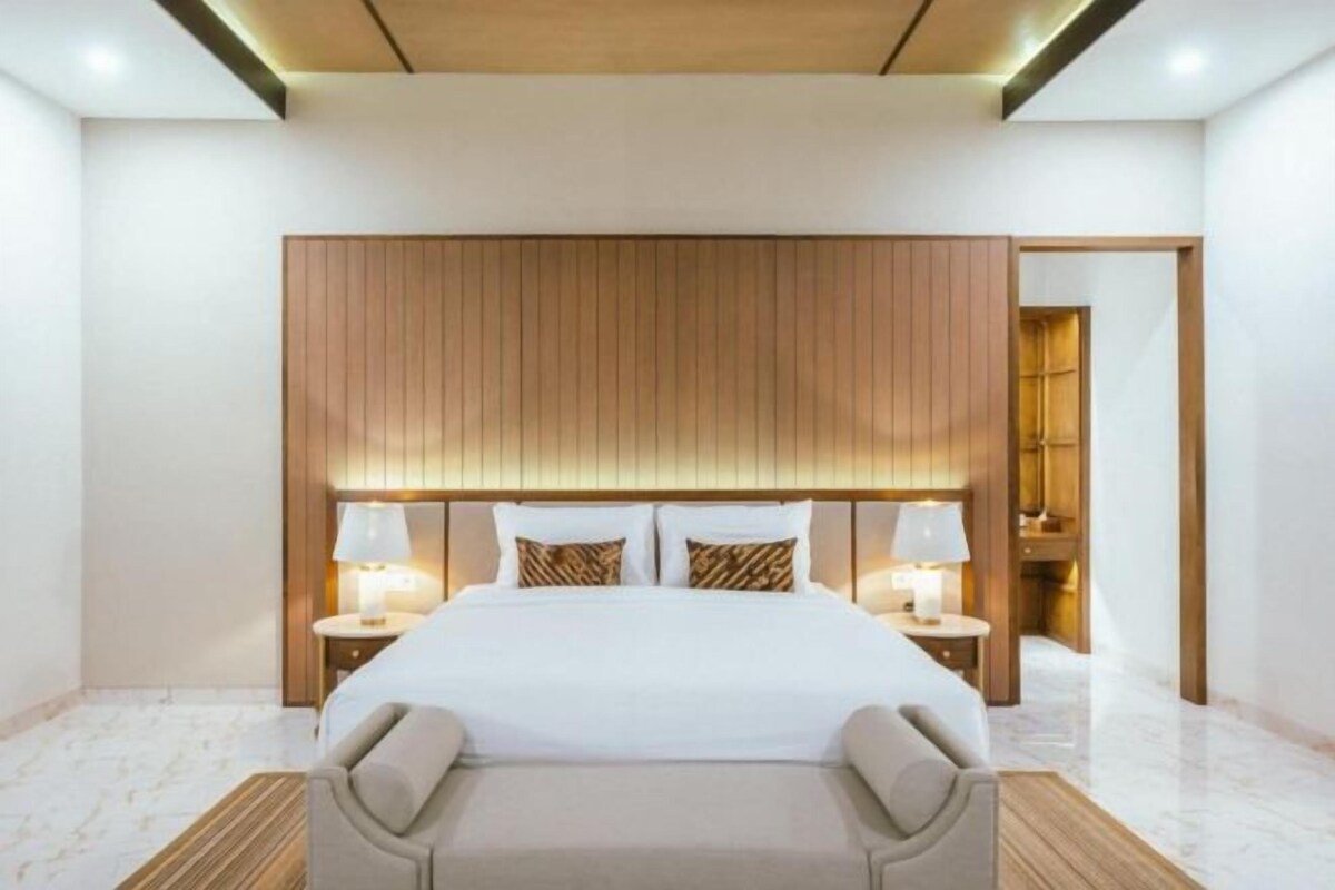 Quarto de hotel moderno com uma cama grande, cabeceira de madeira, duas mesas de cabeceira com abajures e piso de mármore.