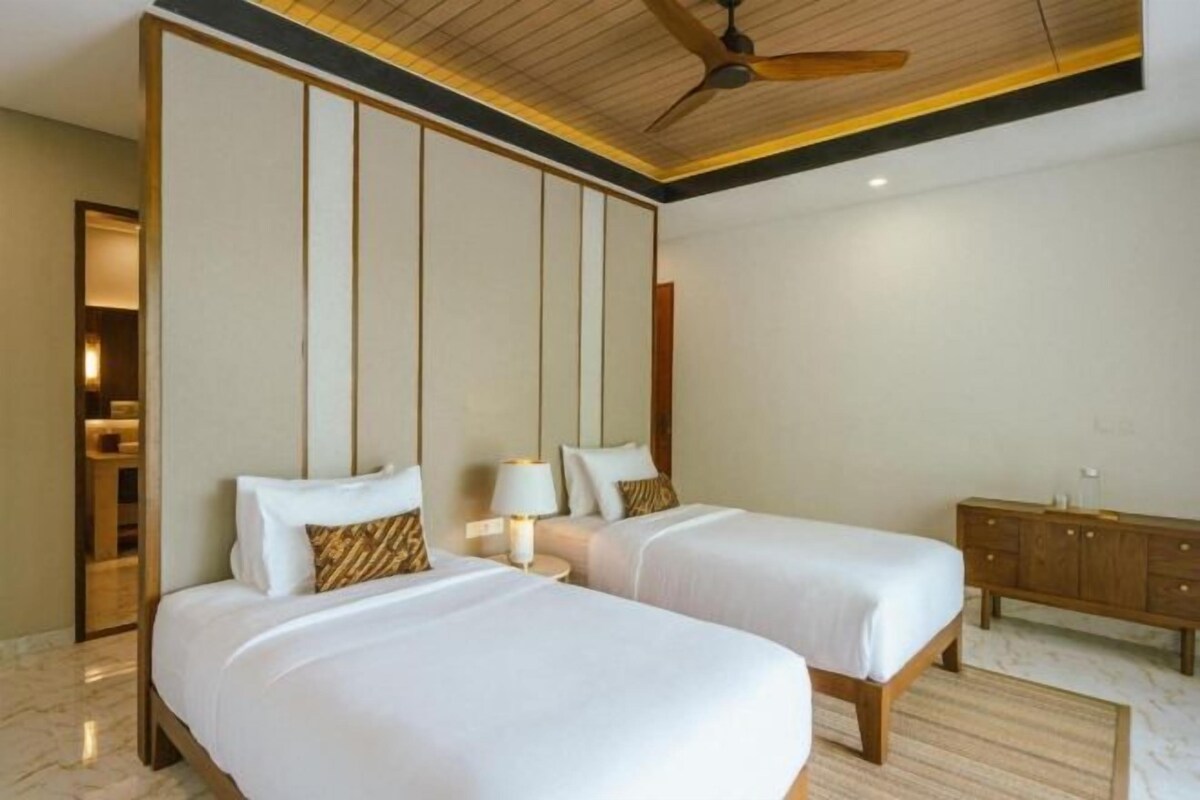 Quarto de hotel com duas camas, um painel de cabeceira de madeira, um ventilador de teto e uma cômoda de madeira.