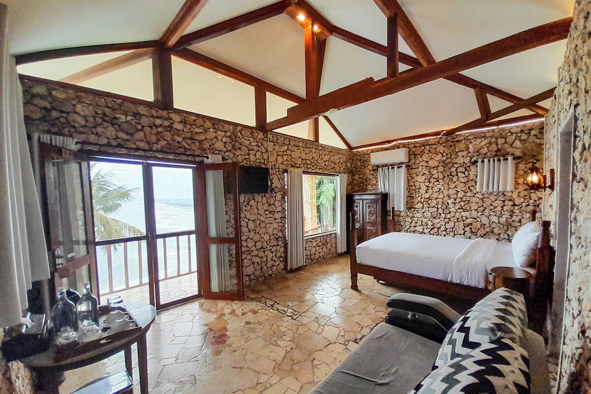 Quarto com parede de pedra, vigas de madeira, cama, sofá e mesa com vista através de uma porta de vidro.