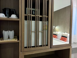 Quarto de hotel com um armário de madeira que contém chávenas e pires brancos, uma máquina de café preta e uma prateleira com chinelos brancos.