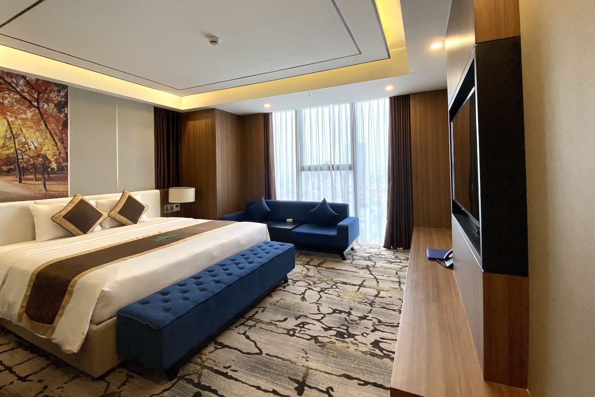 Quarto de hotel moderno com uma cama grande, um sofá azul, uma televisão de tela plana e uma janela ampla com cortinas.