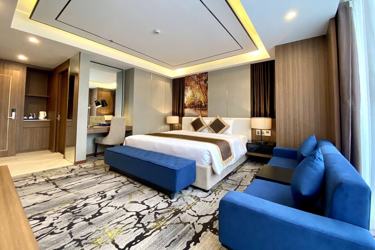 Quarto de hotel moderno com uma cama grande, uma escrivaninha, uma cadeira, um sofá azul e um tapete estampado.