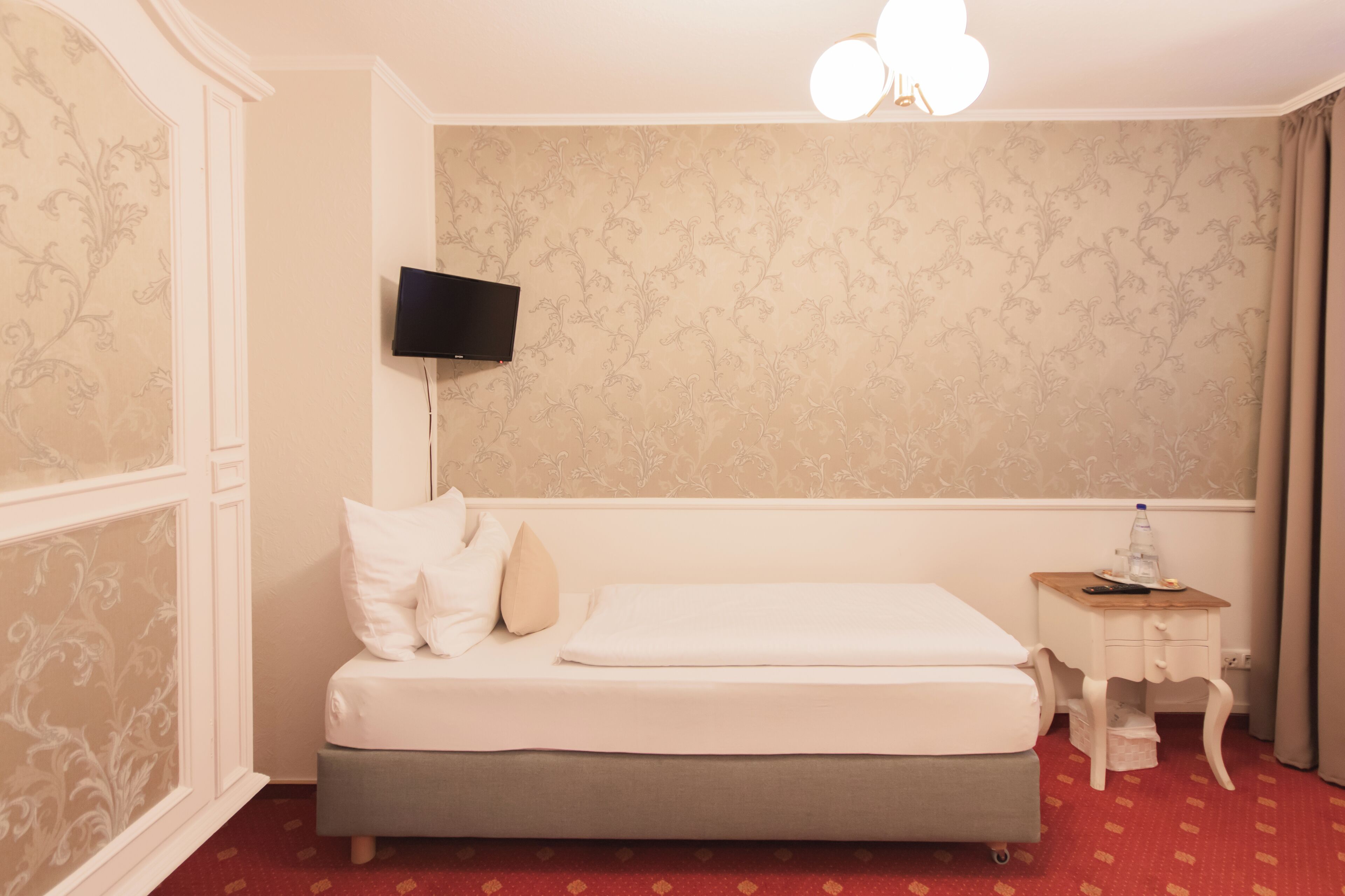 Quarto de hotel com cama, televisão fixada na parede, mesa de cabeceira com garrafa e controle remoto, e tapete vermelho.