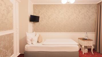 Quarto de hotel com cama, televisão fixada na parede, mesa de cabeceira com garrafa e controle remoto, e tapete vermelho.