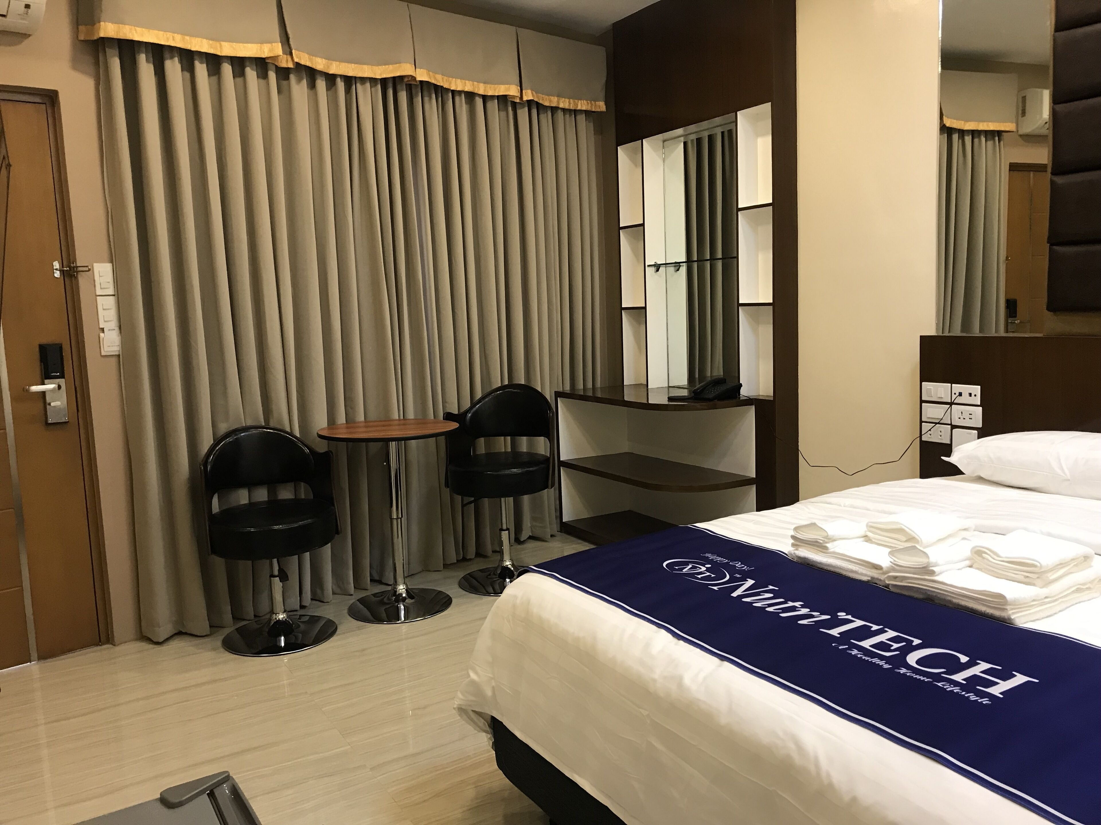 Quarto de hotel com cama, escrivaninha, duas cadeiras, espelho e cortina.