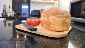 Uma barra de pão sobre uma tábua de corte de madeira com tomates e uma faca em uma bancada de cozinha.