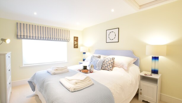 Uma cama bem arrumada com colcha azul e branca, uma mesa de cabeceira com abajur e uma janela com cortina listrada.