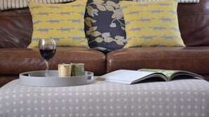 Um sofá de couro marrom com duas almofadas estampadas, um copo de vinho tinto, um tabuleiro com velas e um livro aberto sobre um banquinho listrado.