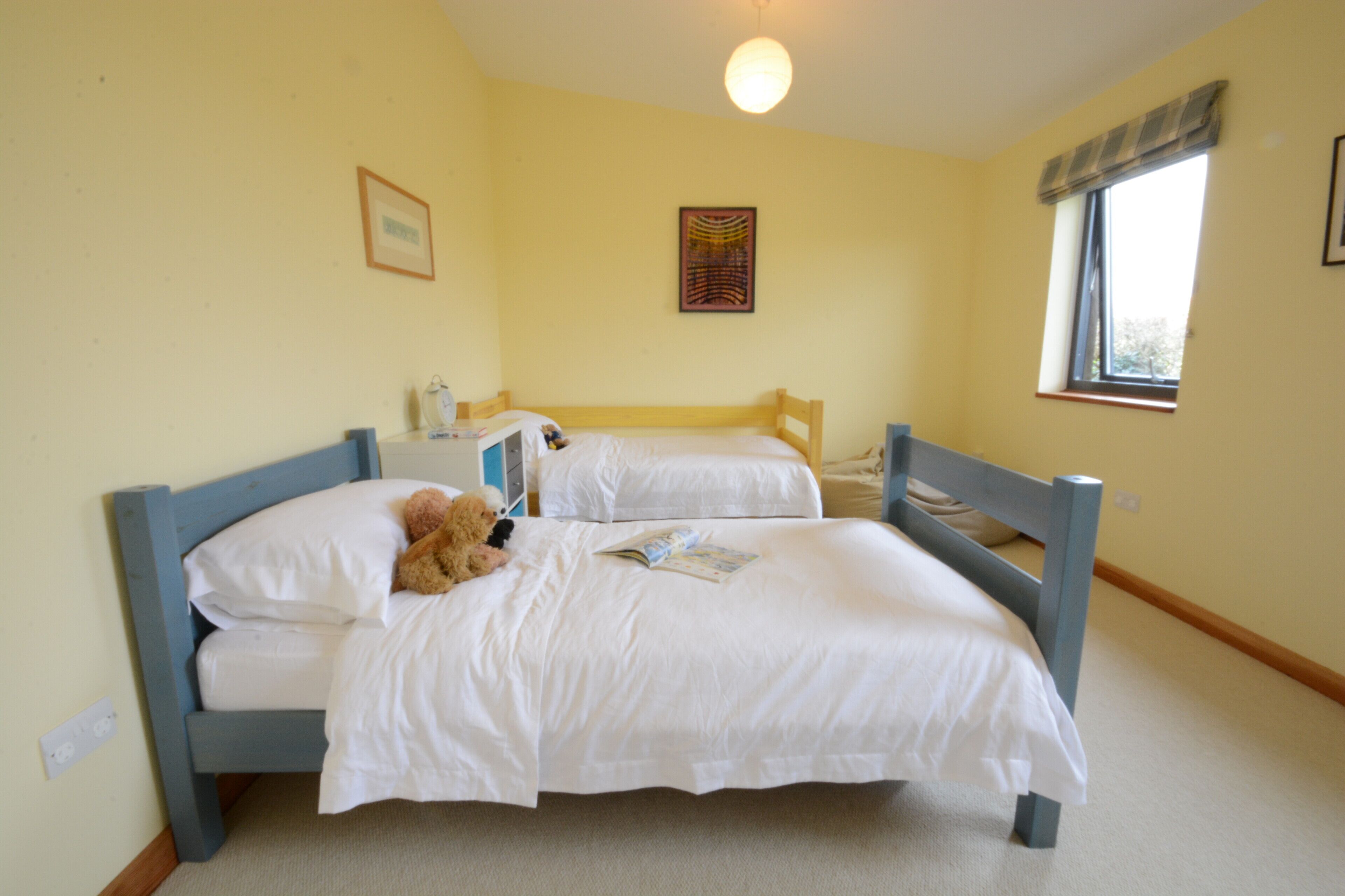 Cama de casal com roupa de cama branca e um urso de pelúcia no lado esquerdo. Há uma janela no lado direito do quarto.