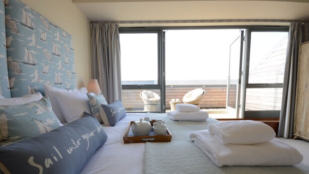 Quarto de hotel com uma cama, um tabuleiro com chá e xícaras, vista para o mar através de uma porta de correr e uma cadeira junto à porta.